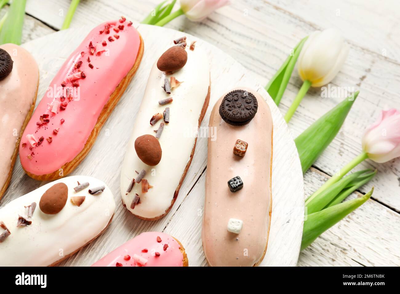 Board with various glazed eclairs and tulips on white wooden background ...