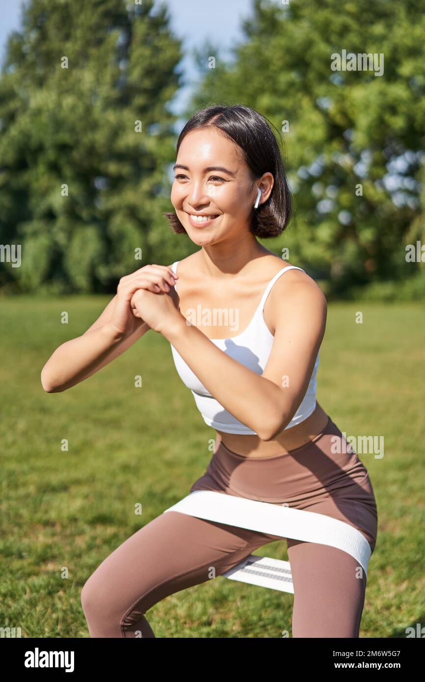 Vertical Shot Of Young Fit Woman Does Squats In Park Using Stretching Band On Legs Smiling