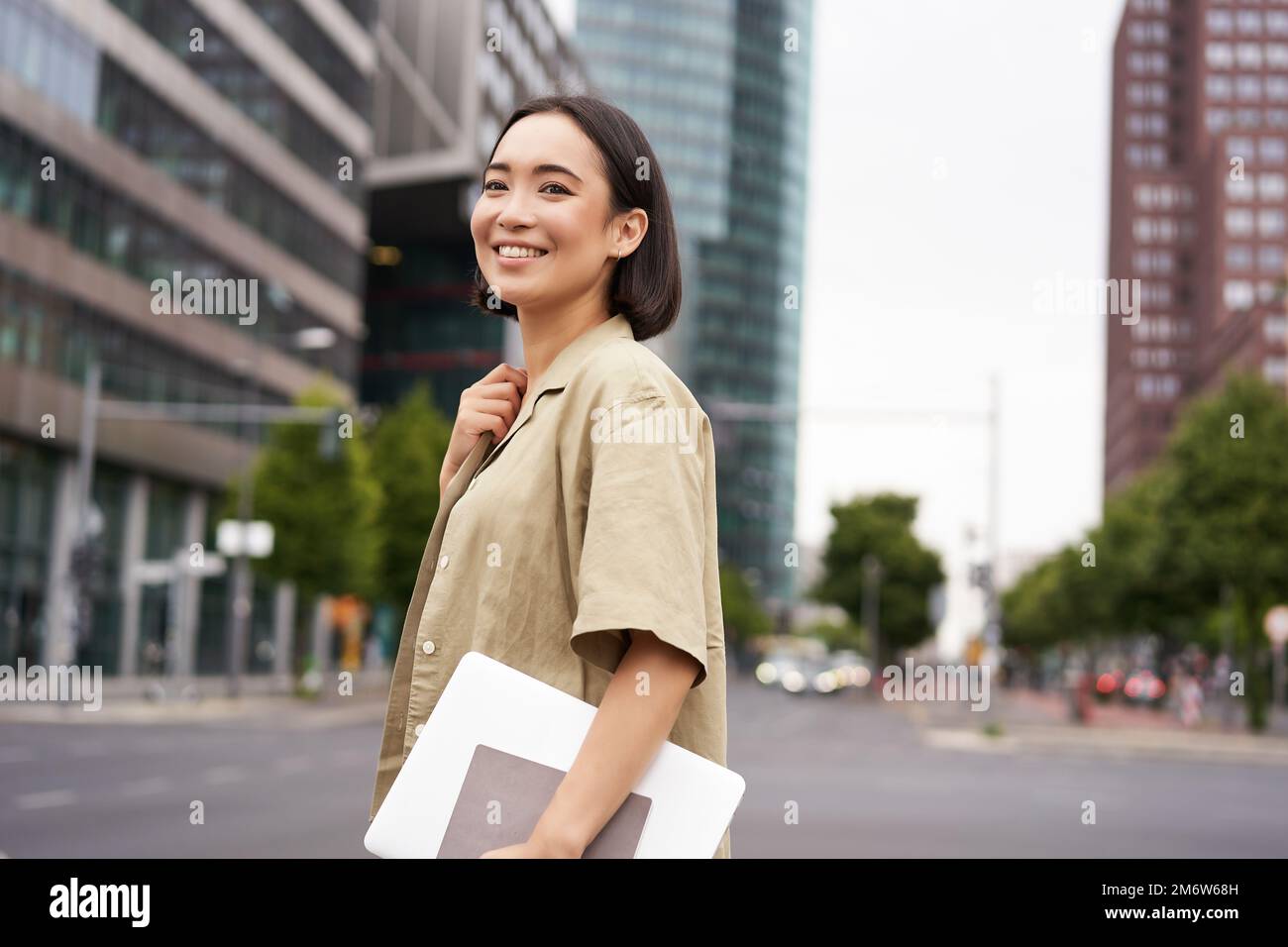 Outdoor shot of asian girl with laptop, going somewhere in city centre, walking on street, going ...