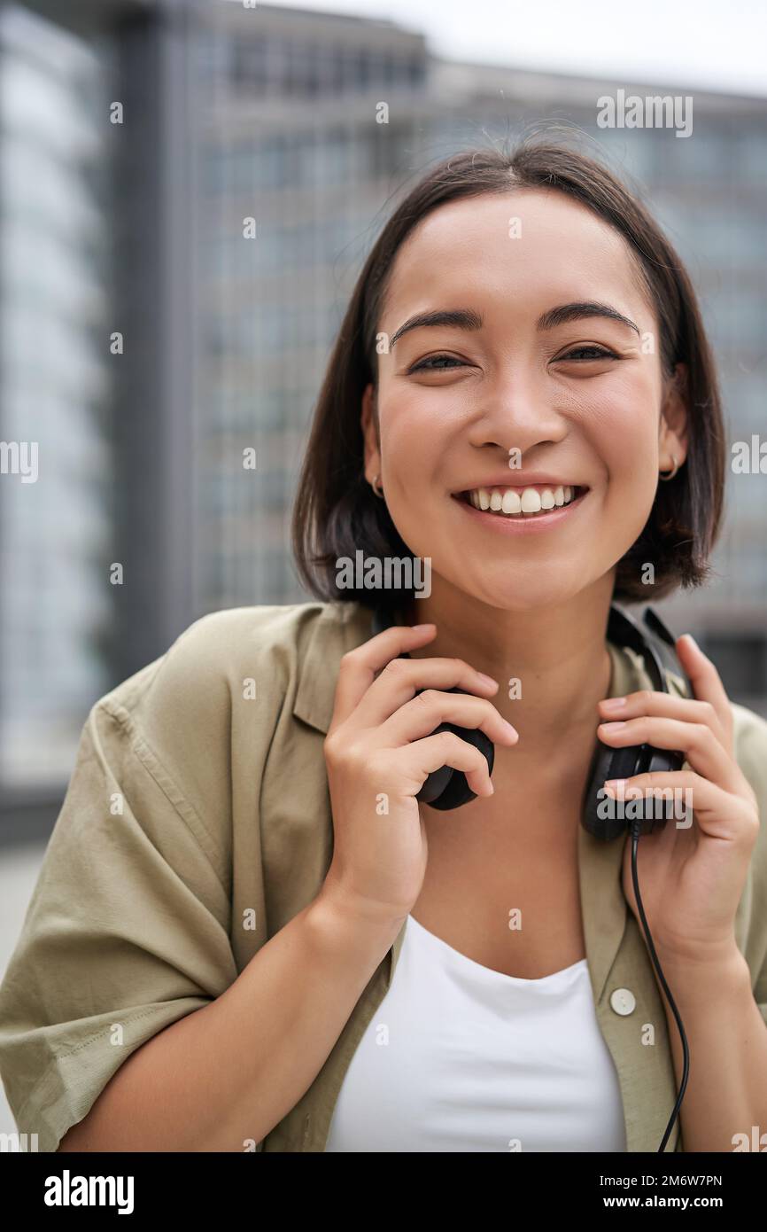 Vertical shot of beautiful asian woman posing with headphones around neck, smiling and laughing ...