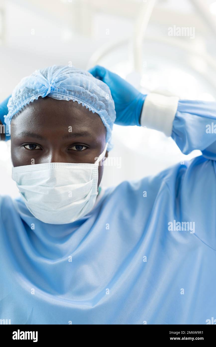 Vertical of african american male surgeon in gown and cap tying on mask ...
