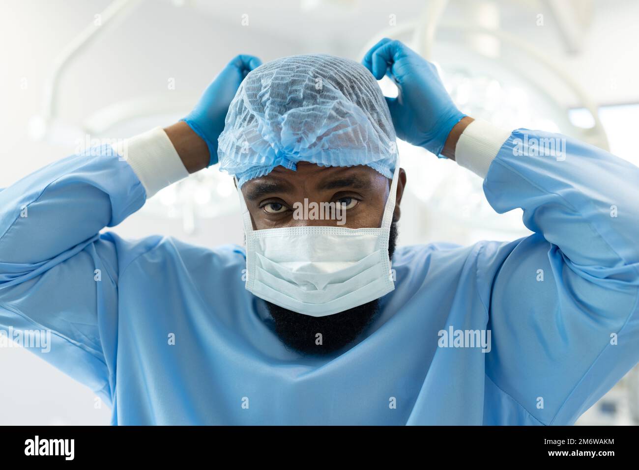 African american male surgeon in gown and cap tying on mask in ...