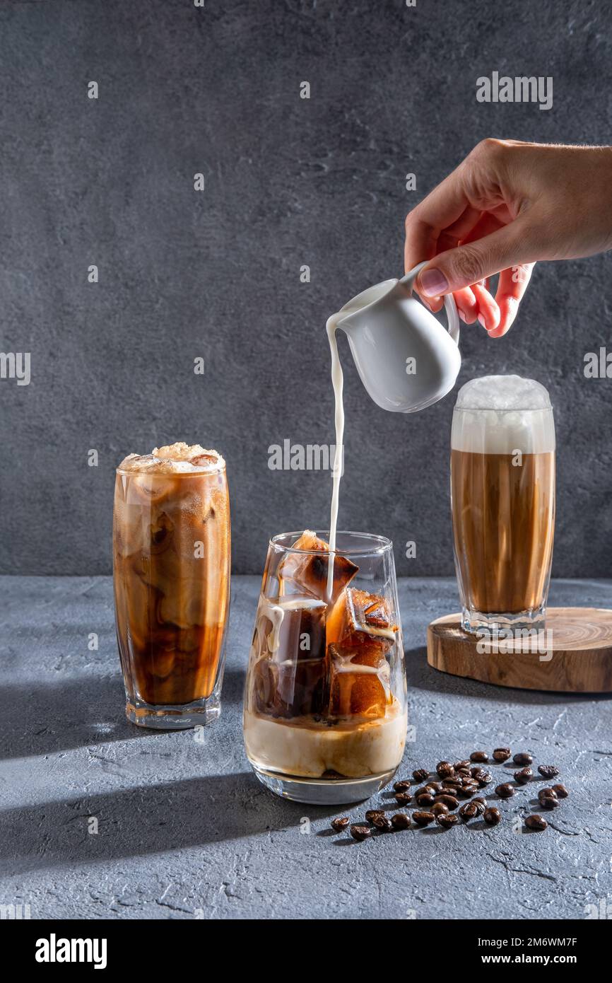 Milk Being Poured Into Iced Coffee on a dark table. Ice coffee in a tall glass with cream poured ...
