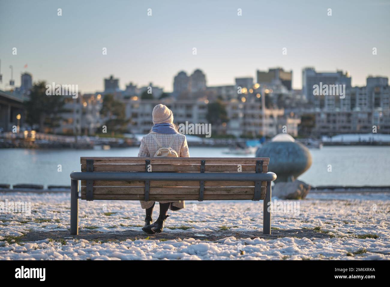 A female seen from behind, sitting on a bench against a lake in a park ...