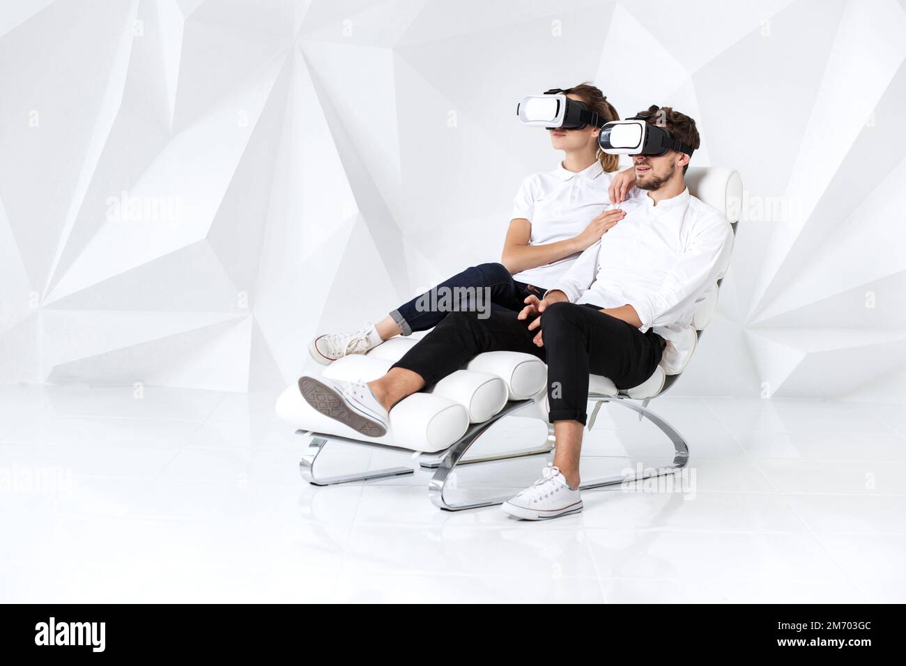 A young couple wearing VR headsets sitting on white chair in a room ...