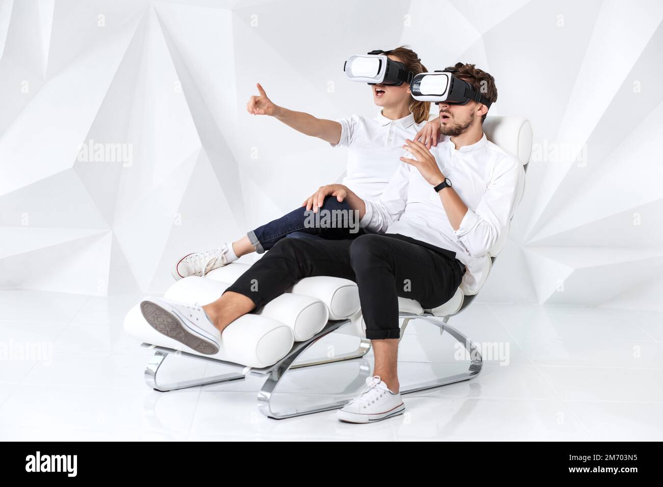 A young couple wearing VR headsets sitting on white chair in a room ...