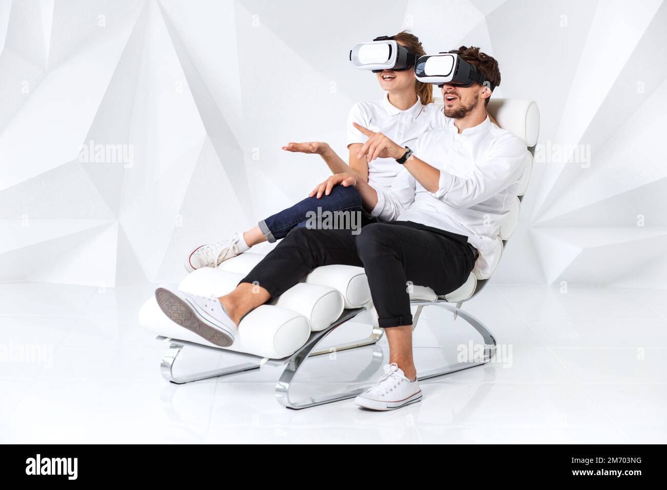 A young couple wearing VR headsets sitting on white chair in a room ...