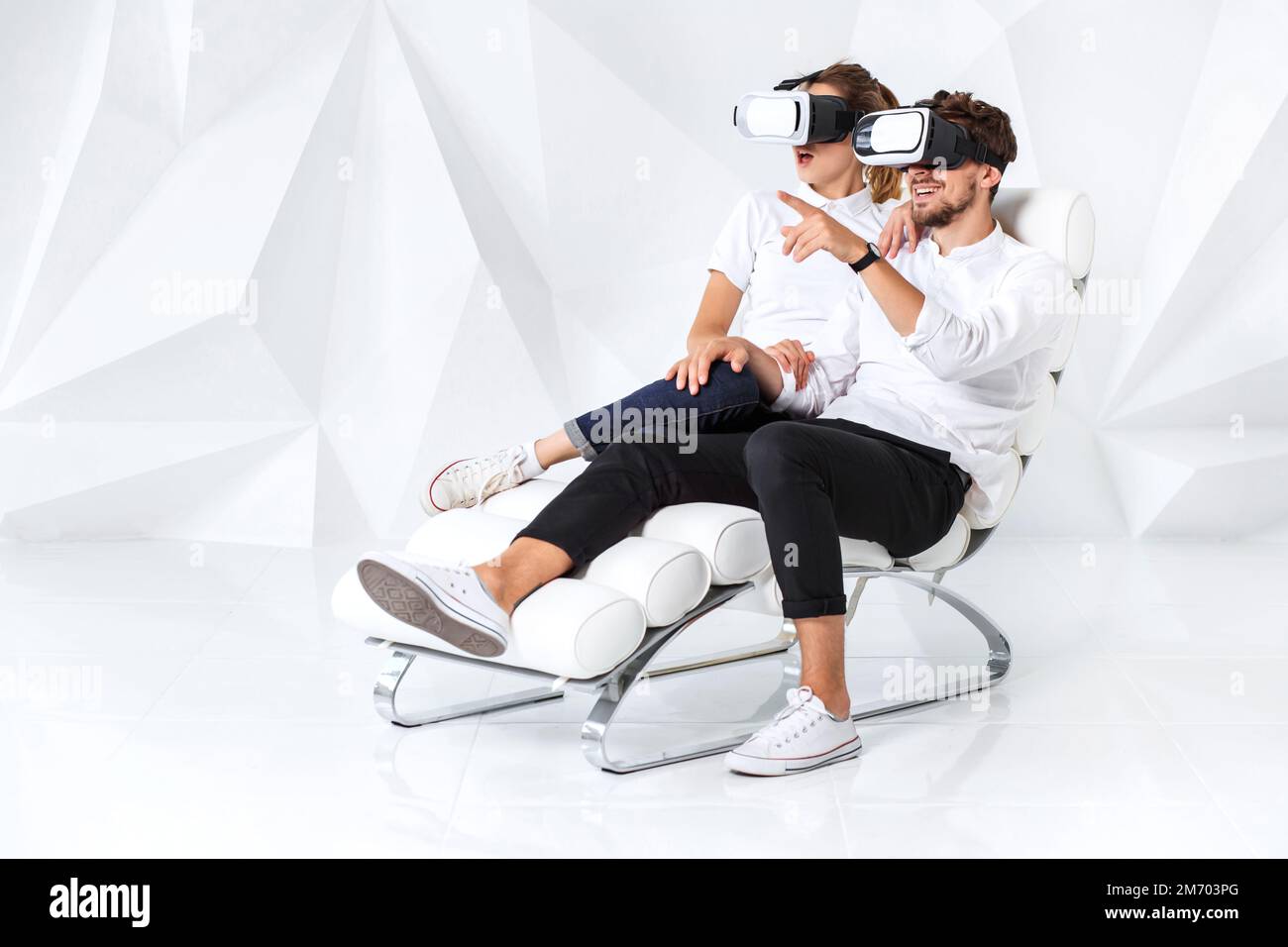 A young couple wearing VR headsets sitting on white chair in a room ...