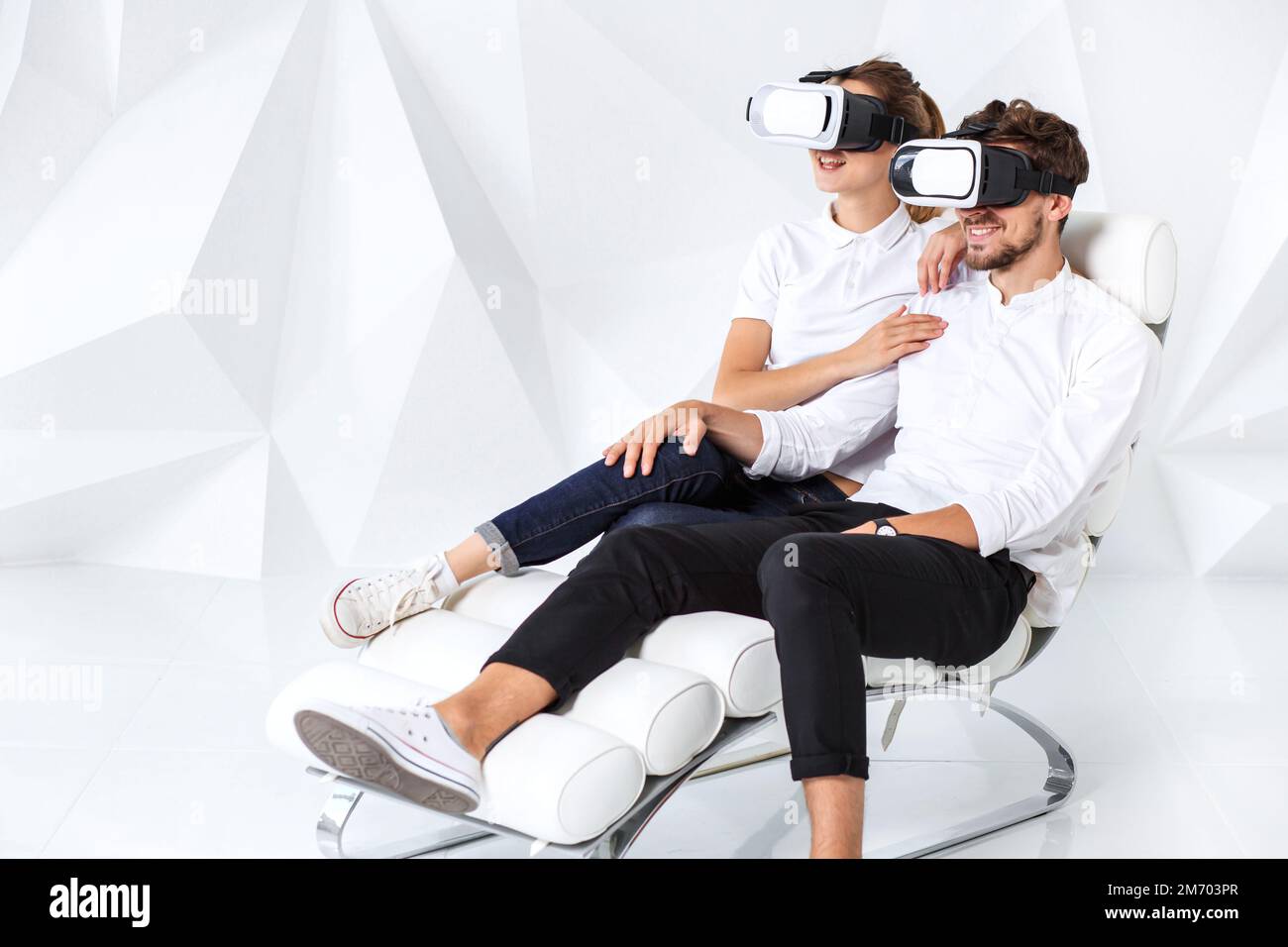 A young couple wearing VR headsets sitting on white chair in a room ...