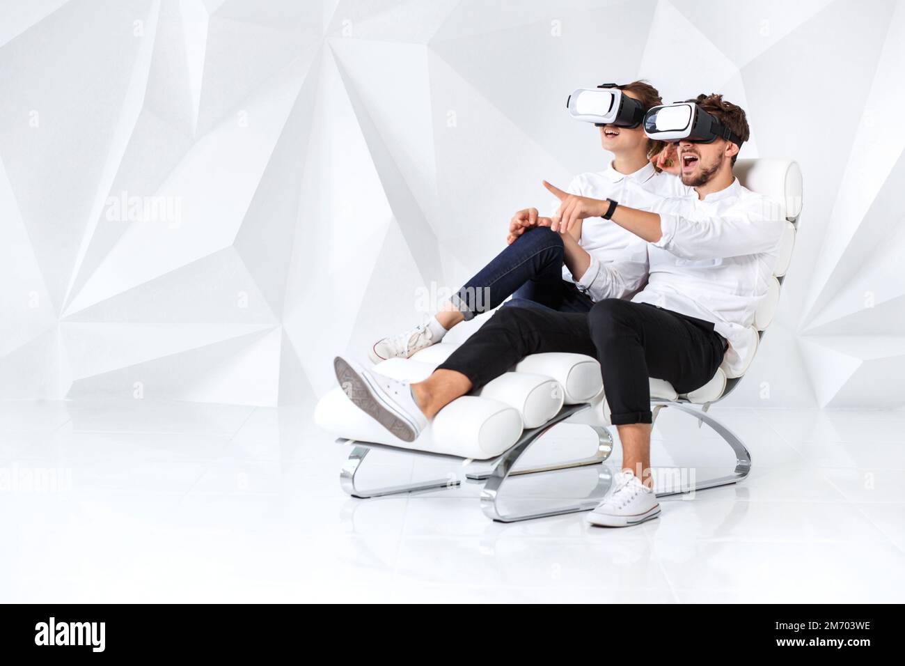 A young couple wearing VR headsets sitting on white chair in a room ...
