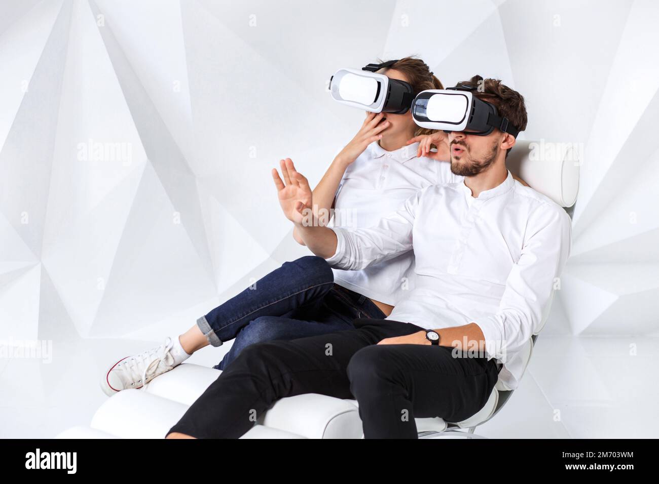 A young couple wearing VR headsets sitting on white chair in a room ...