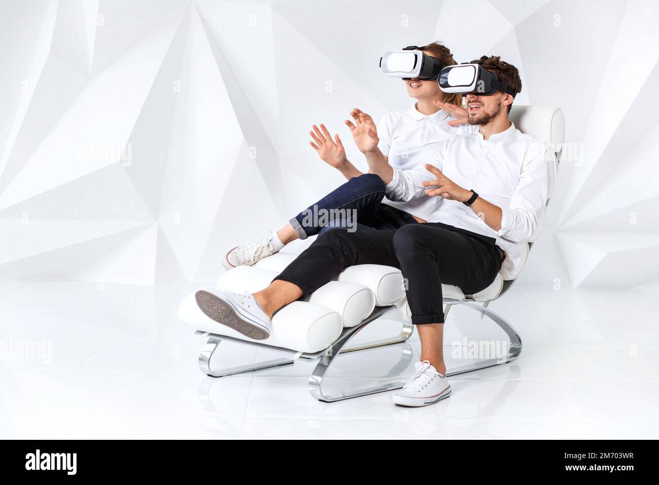 A young couple wearing VR headsets sitting on white chair in a room ...