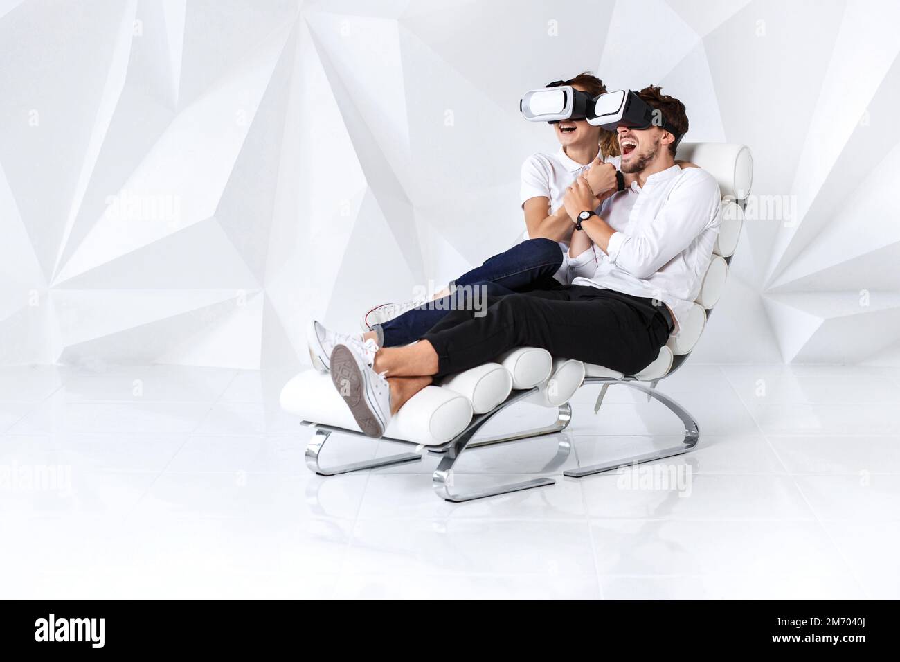 A young couple wearing VR headsets sitting on white chair in a room ...