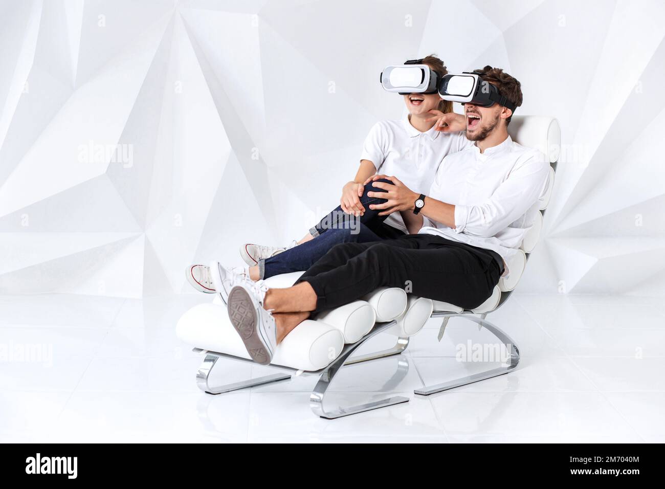 A young couple wearing VR headsets sitting on white chair in a room ...