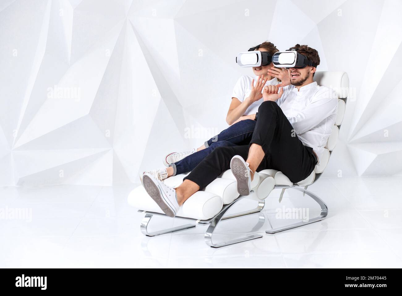 A young couple wearing VR headsets sitting on white chair in a room ...