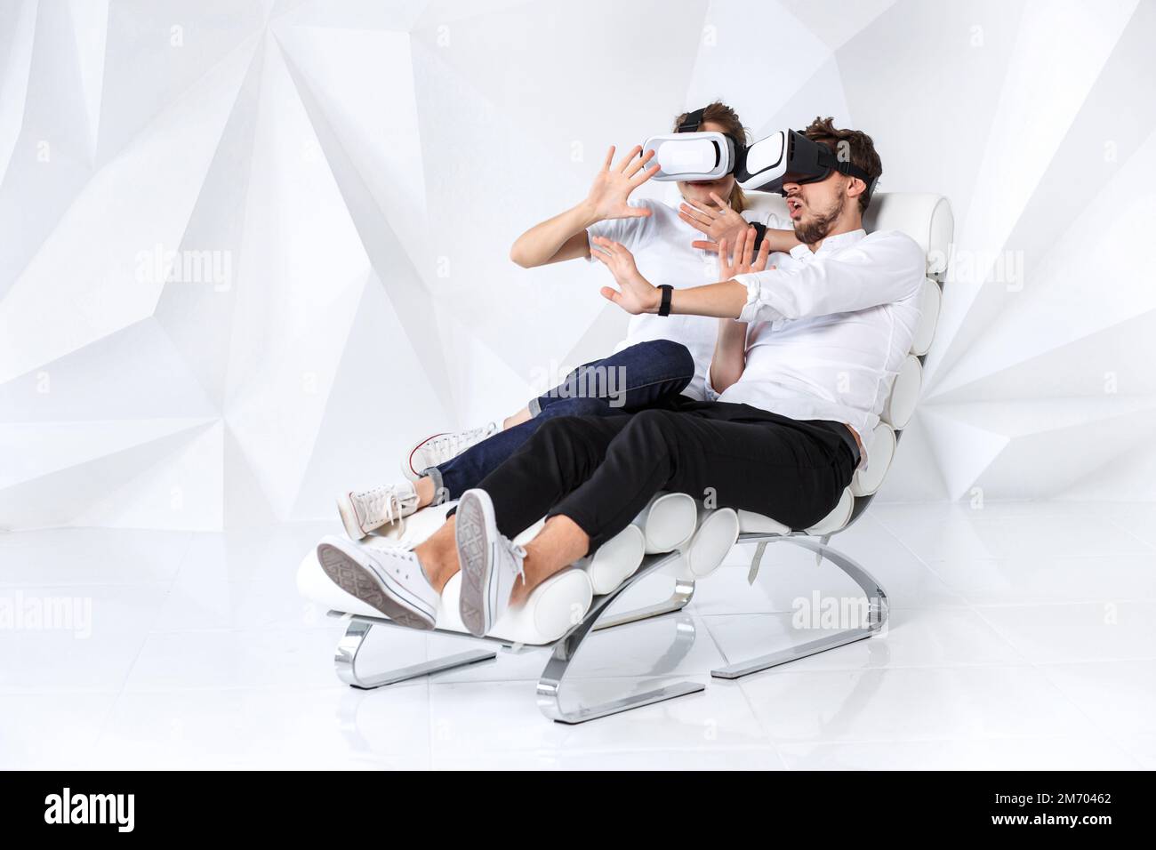 A young couple wearing VR headsets sitting on white chair in a room ...