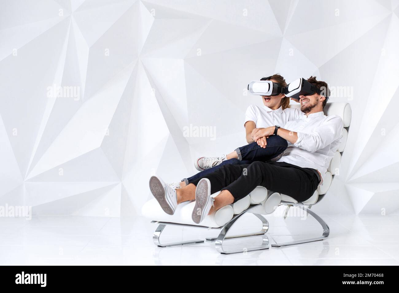 A young couple wearing VR headsets sitting on white chair in a room ...