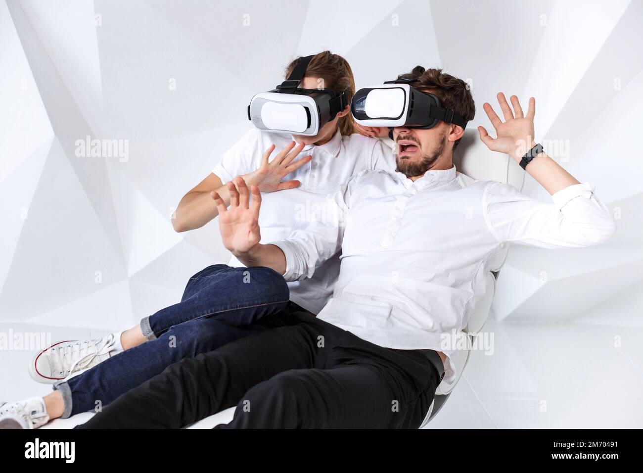 A young couple wearing VR headsets sitting on white chair in a room ...