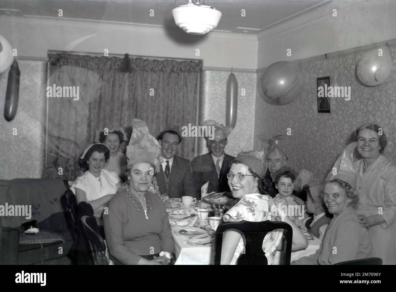 1950s, historical, family party, big smiles as a family, most wearing party hats, sit in a front ...