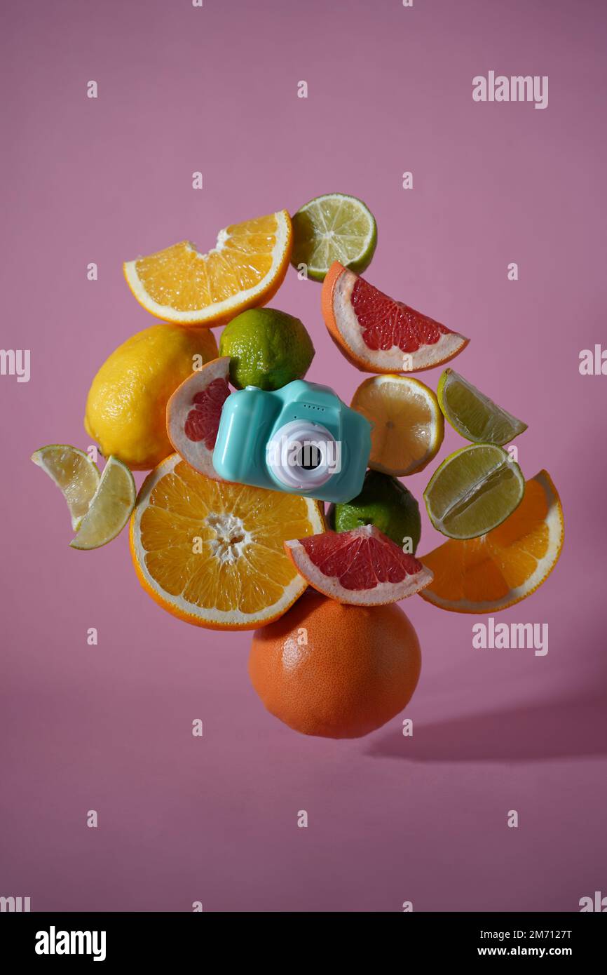 Whole and cut fruits and toy camera float in the air on a pink ...