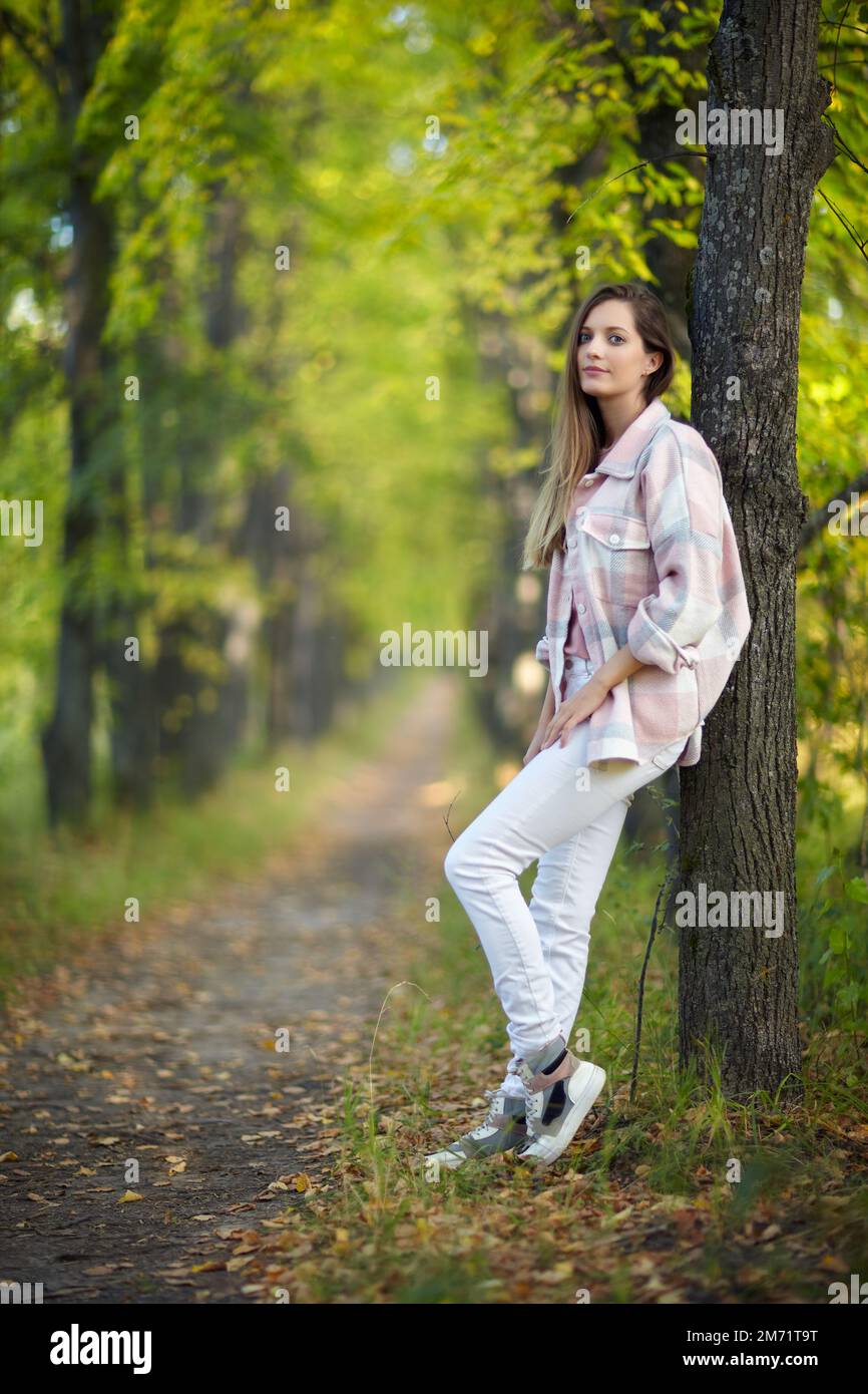 Blonde female in blue dress leaning against a tree trunk in autumn ...