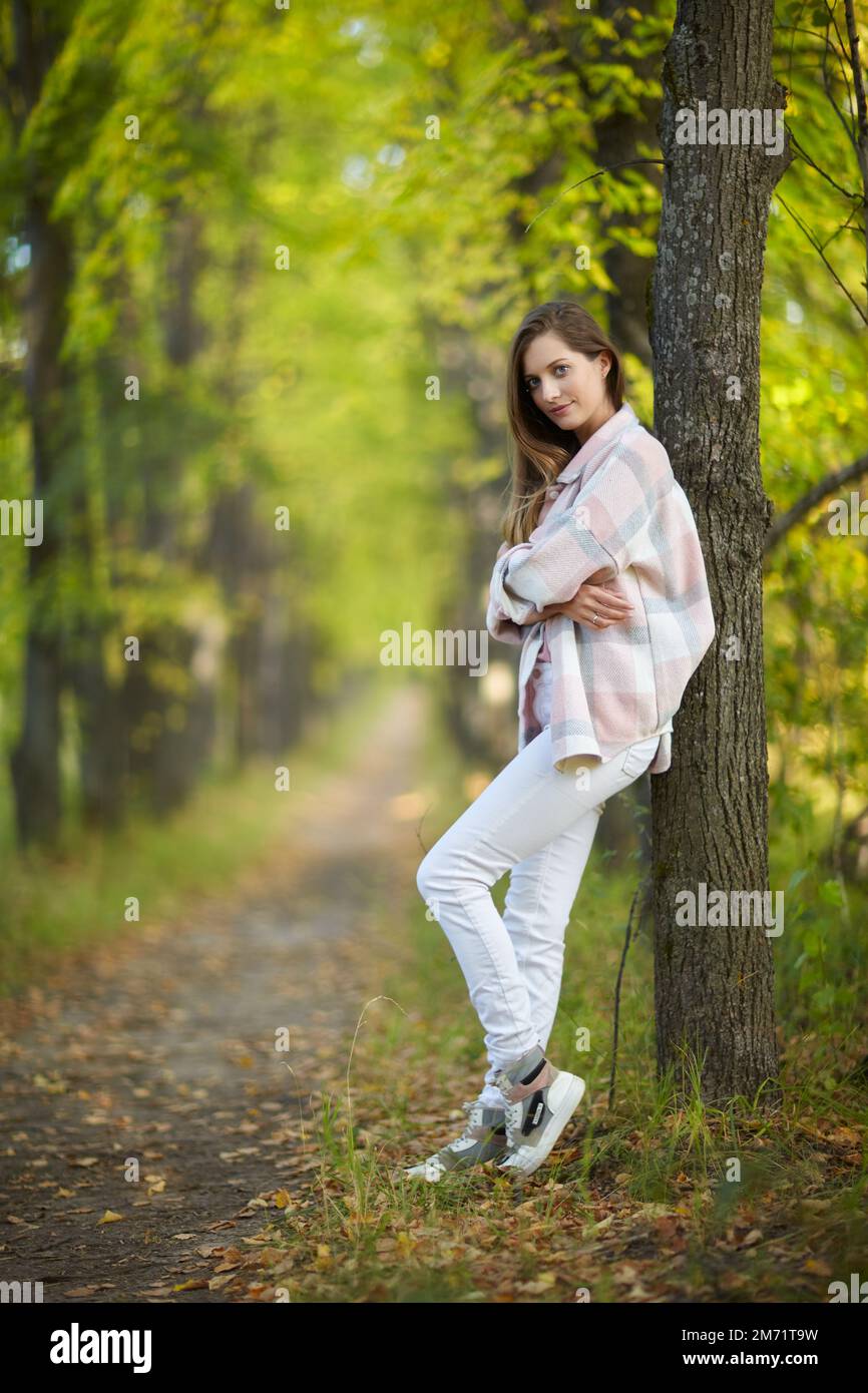 Blonde female in blue dress leaning against a tree trunk in autumn ...