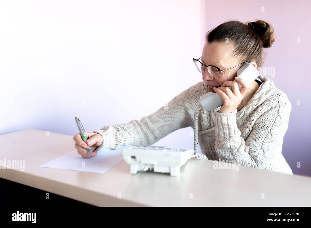 Middle-aged woman talks on phone and writting notes at her workplace ...
