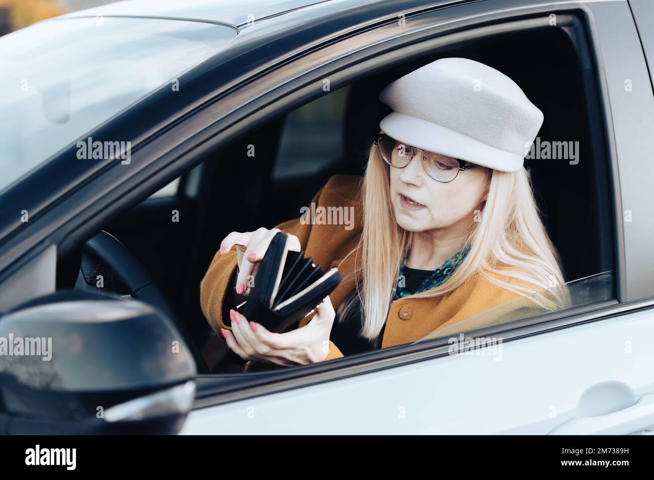 Middle-aged woman sits behind the wheel of car and looks for money in ...