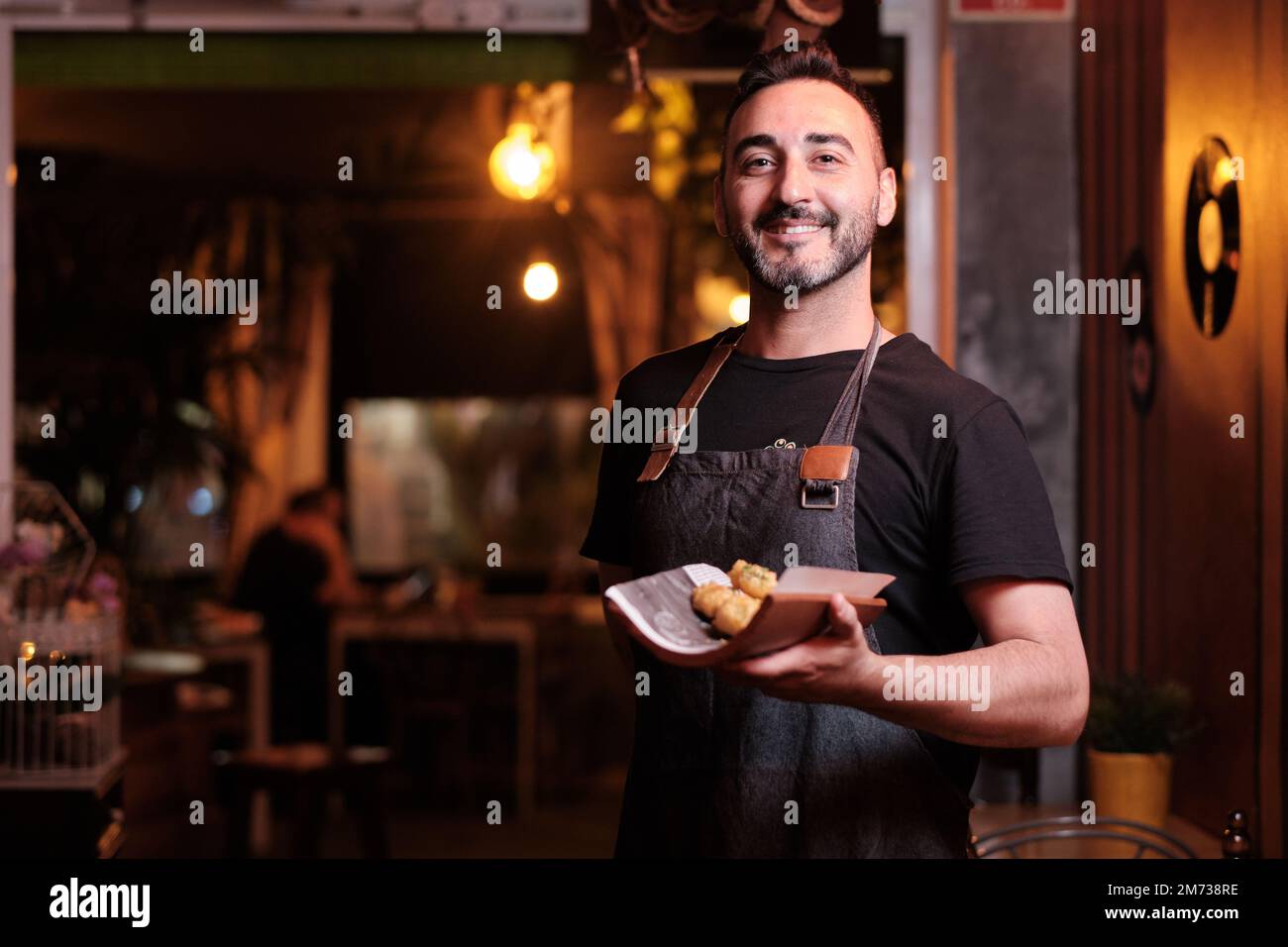 Male waiter in uniform smiling while holding a plate with food in a ...