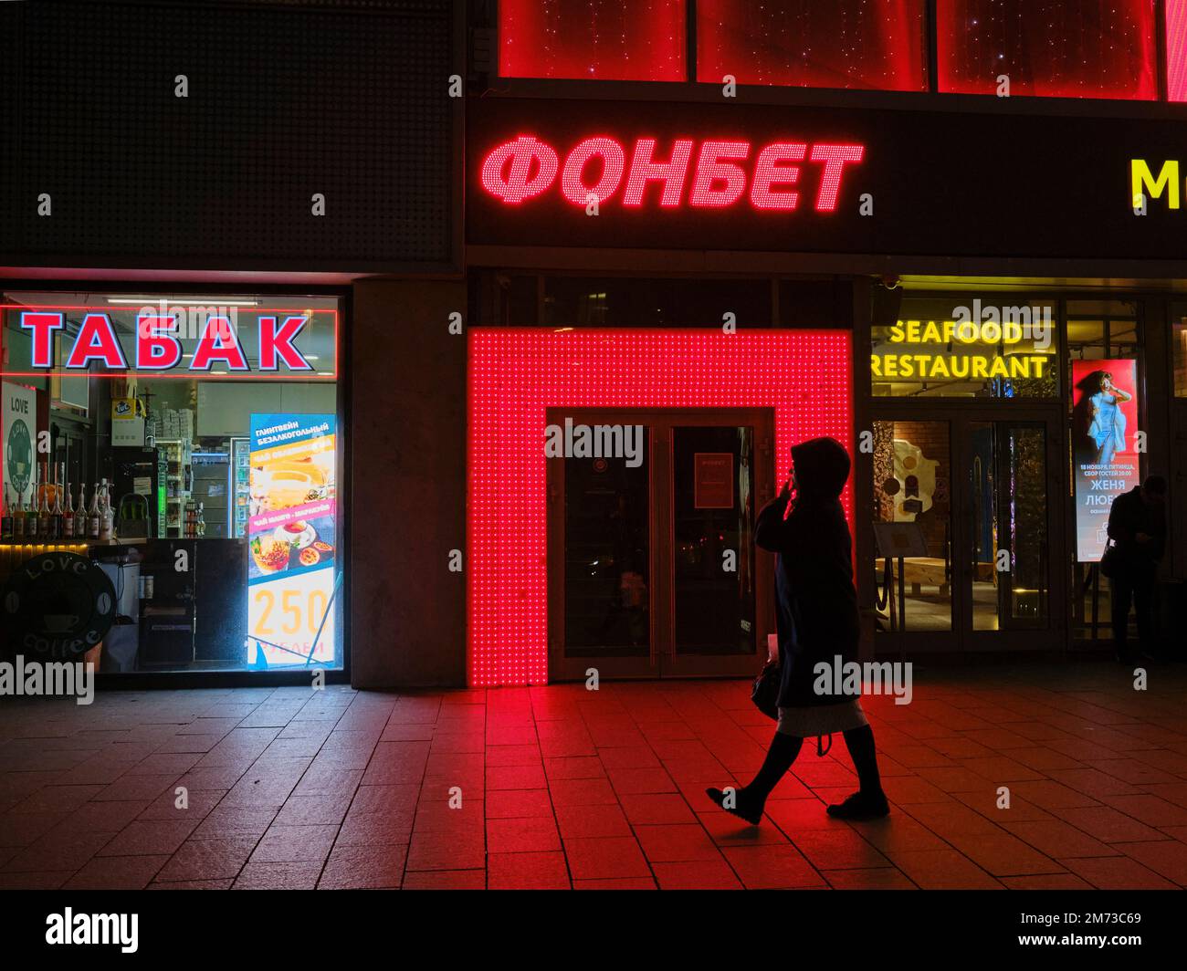 Woman walking along the sidewalk passes by shops' windows illuminated ...