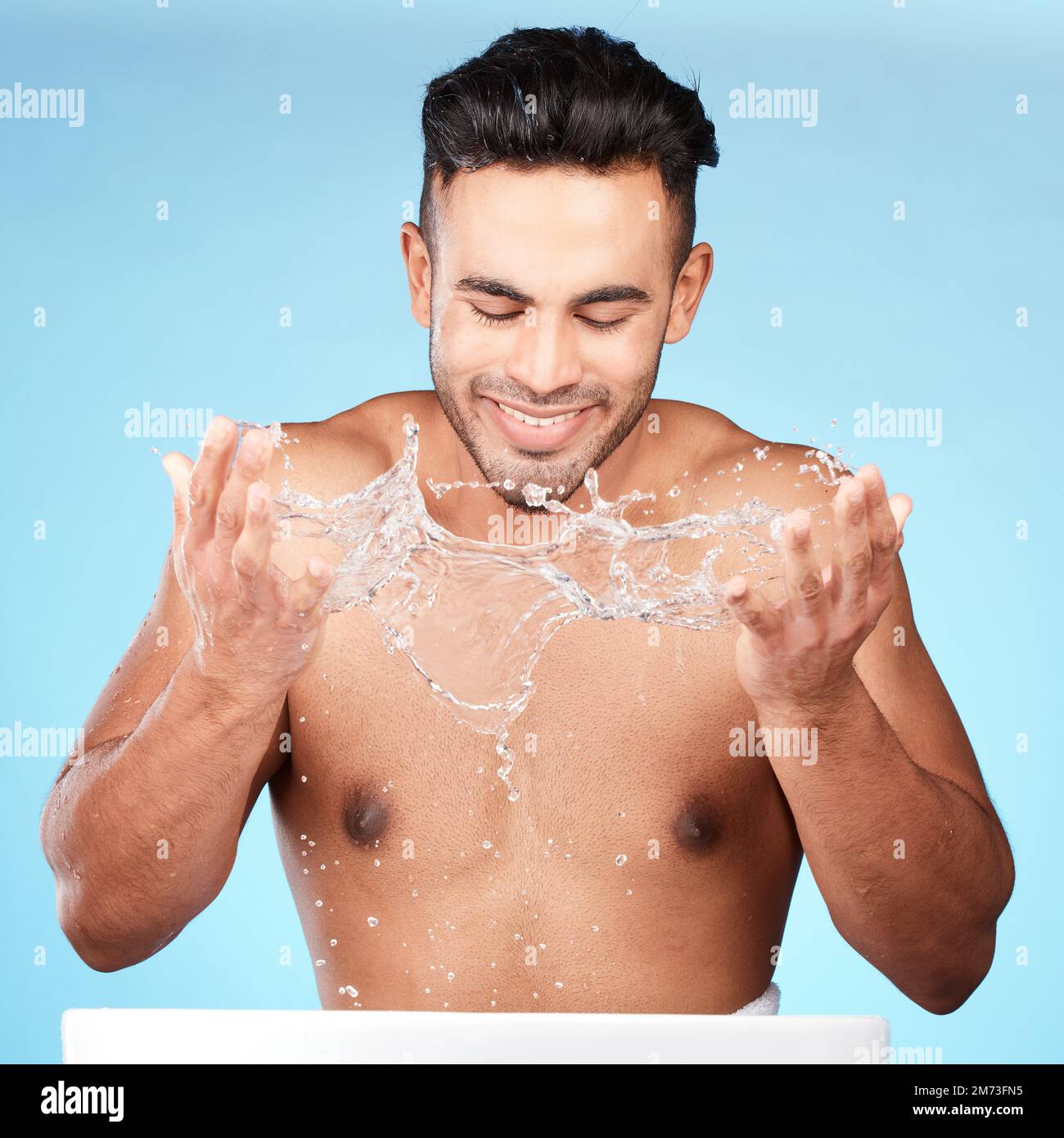 Face, water splash and skincare of man cleaning in studio isolated on a blue background. Hygiene ...