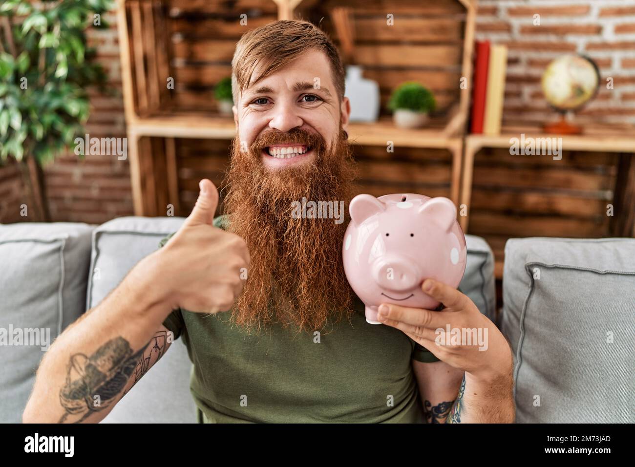 Redhead man holding piggy bank smiling happy and positive, thumb up ...