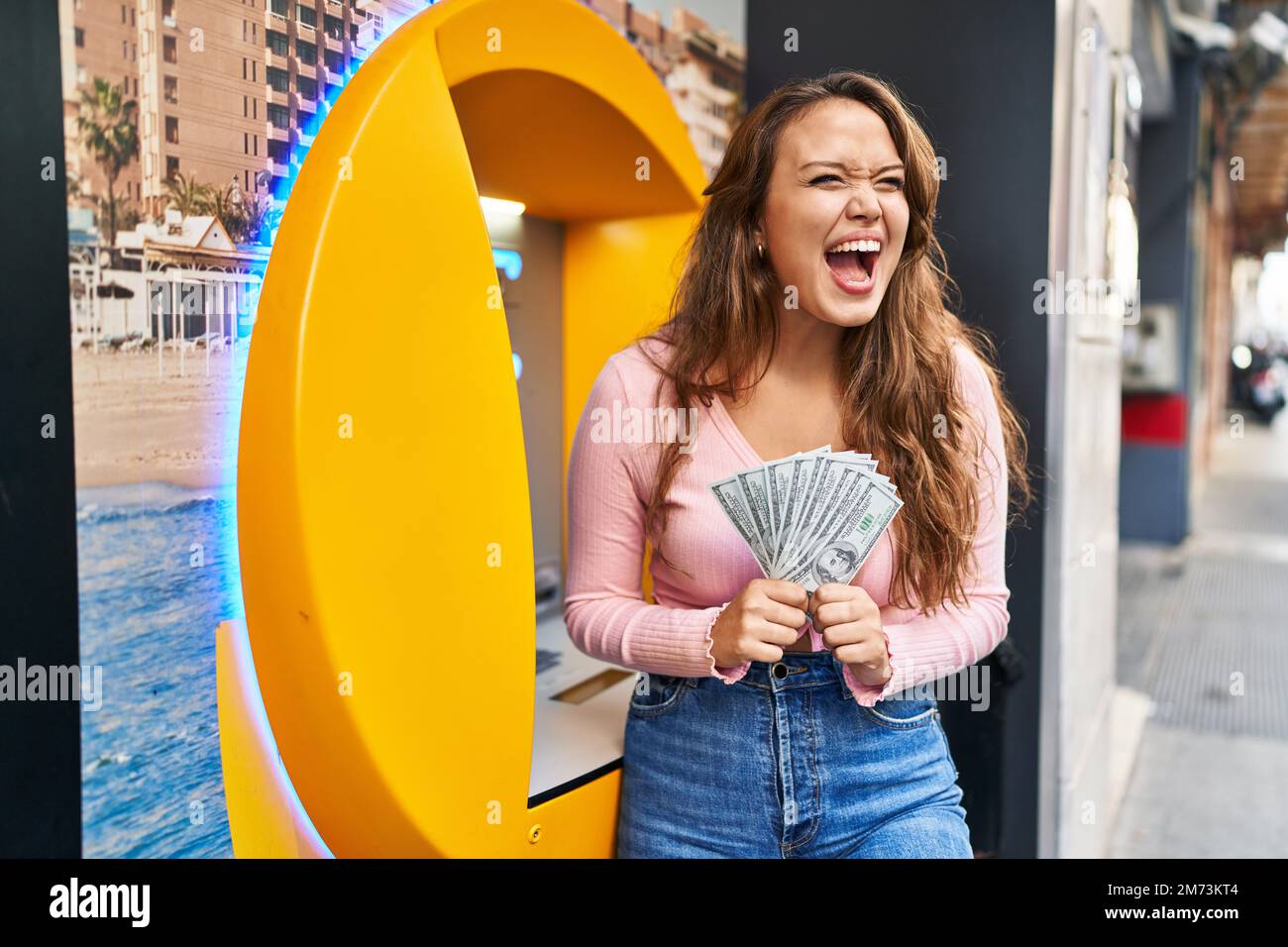 Young hispanic woman taking cash from atm angry and mad screaming ...
