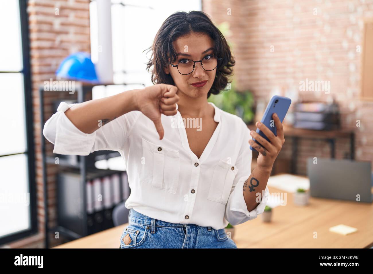 Young hispanic woman working at the office using smartphone with angry ...