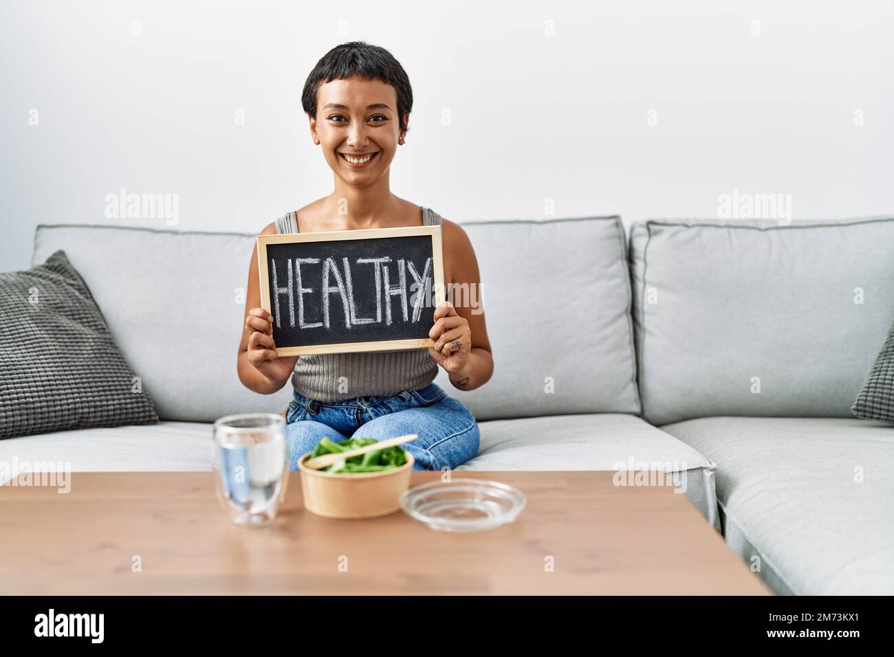 Young hispanic woman eating salad holding healthy blackboard at home ...