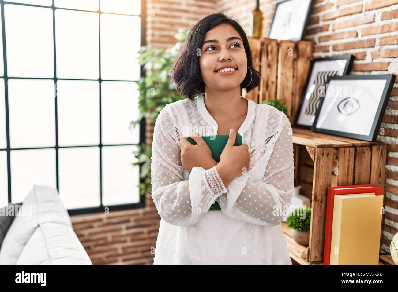 Young latin woman smiling confident hugging book at home Stock Photo ...