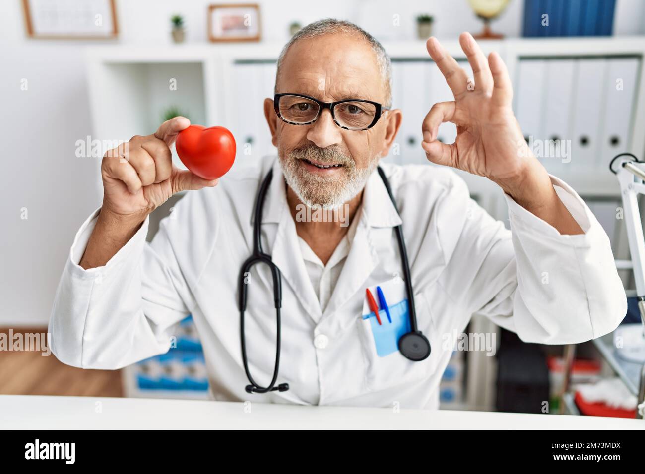 Mature doctor man holding red heart at the clinic doing ok sign with ...