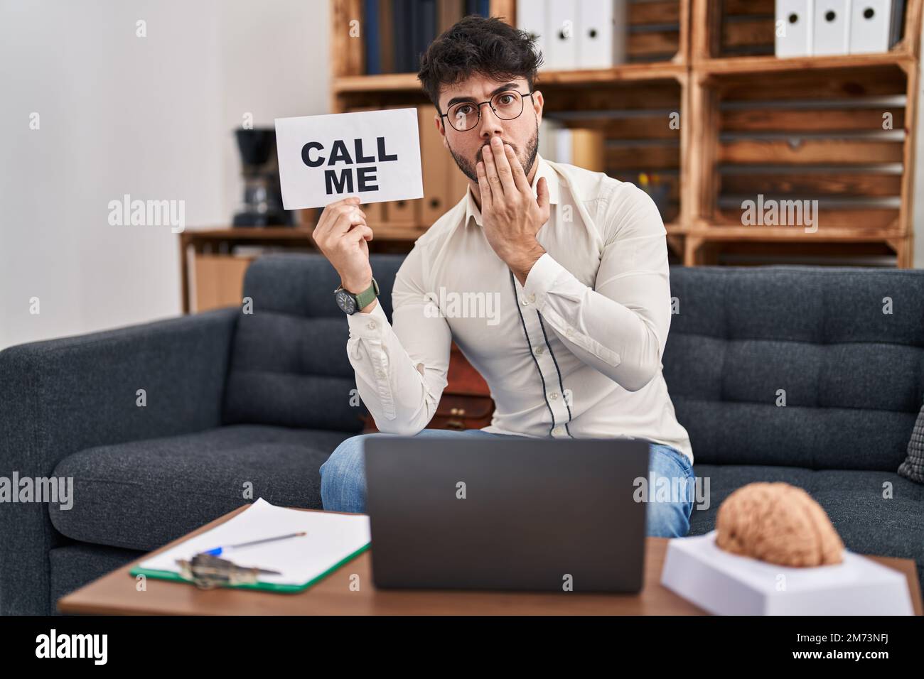 Hispanic man with beard working at therapy office holding call me sign ...
