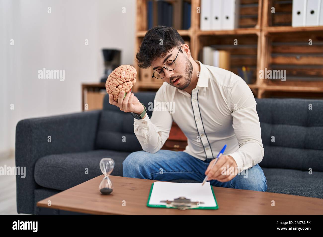 Young hispanic man psychologist writing on document holding brain ...