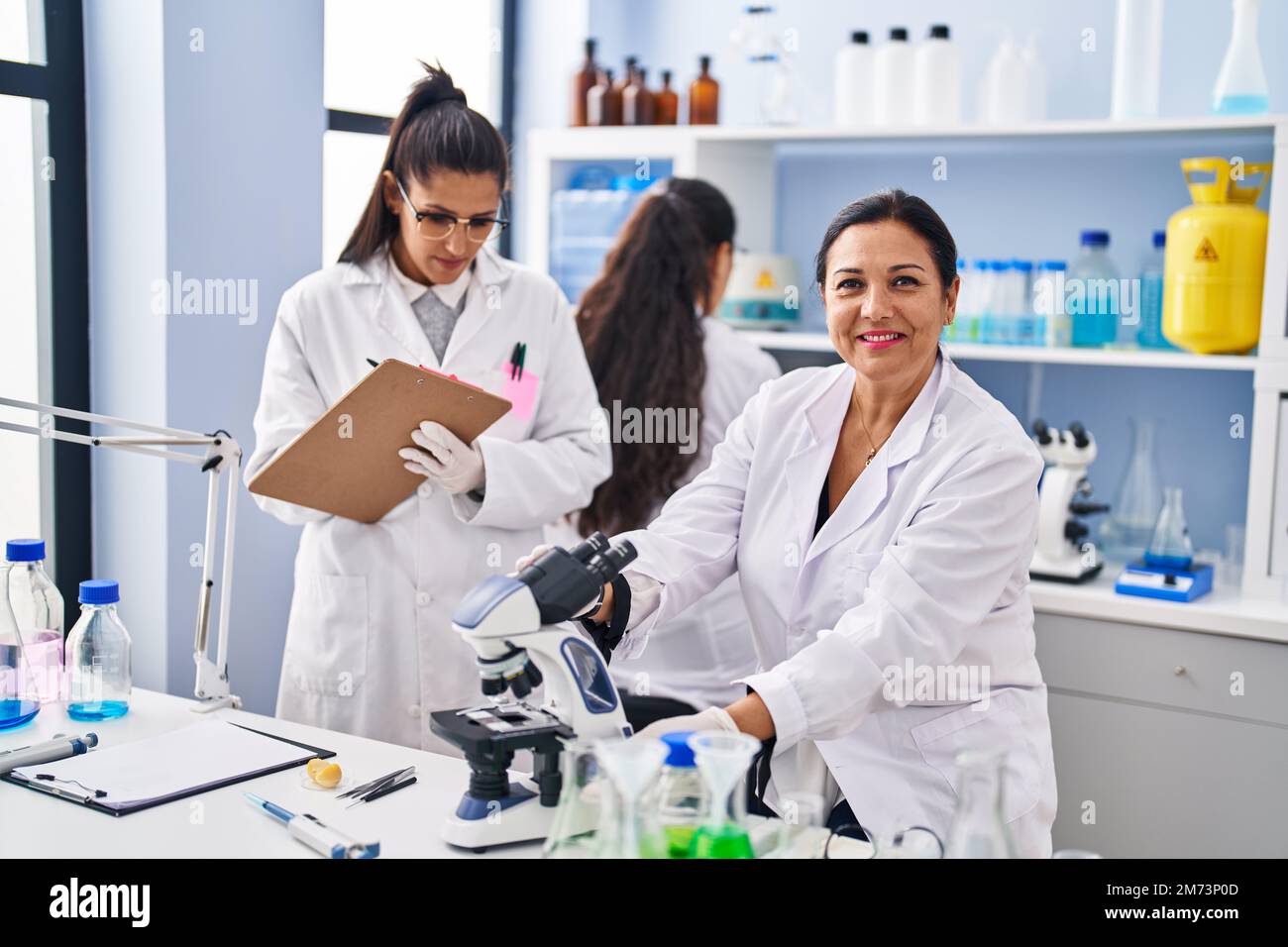 Three woman scientists using microscope write on checklist at ...