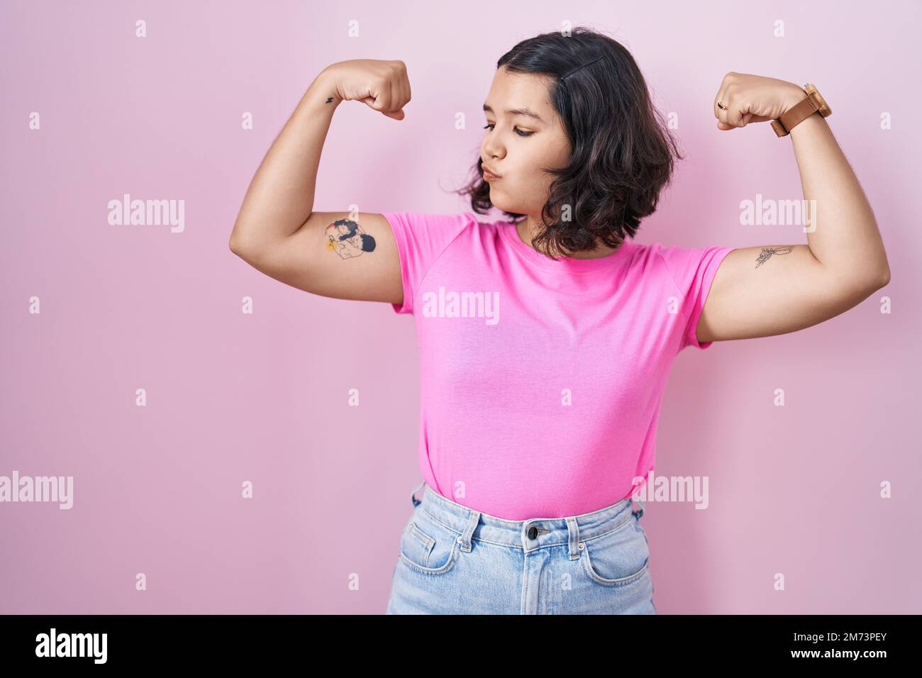 Young hispanic woman standing over pink background showing arms muscles ...