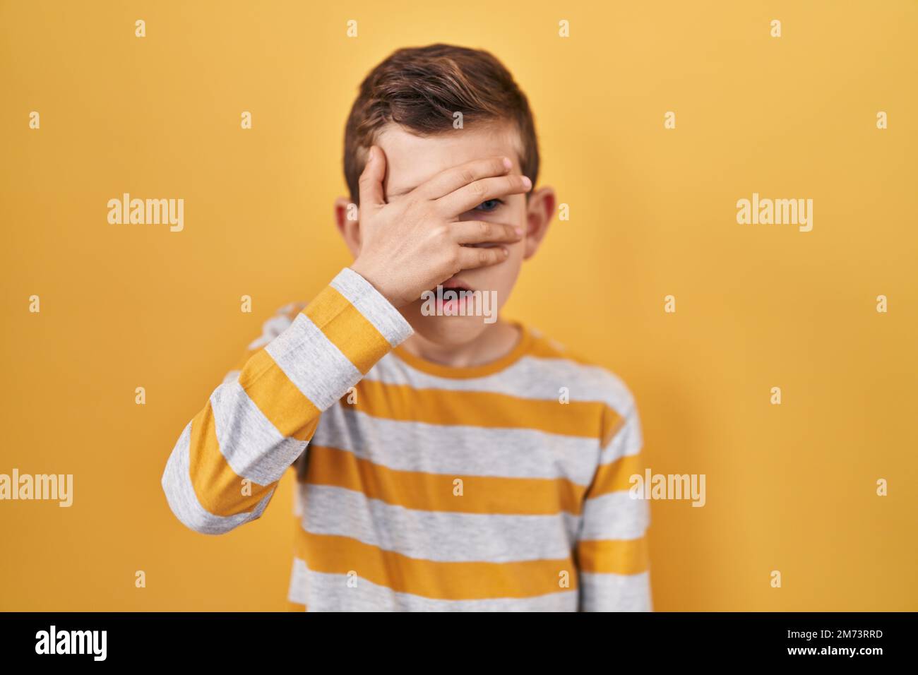Young caucasian kid standing over yellow background peeking in shock ...