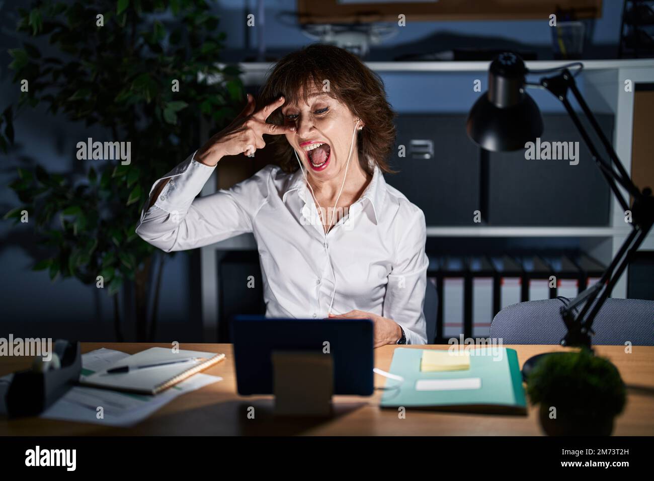 Middle age woman working at the office at night doing peace symbol with ...