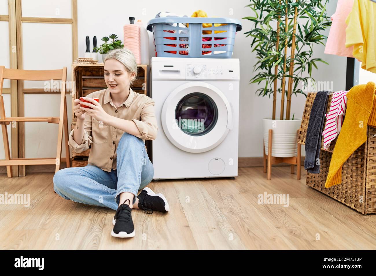 Young caucasian woman drinking coffee waiting for washing machine at ...