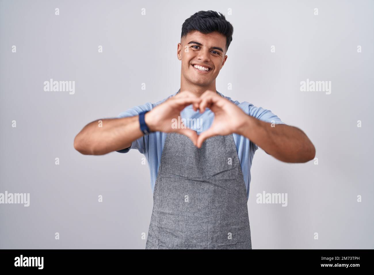 Hispanic young man wearing apron over white background smiling in love ...