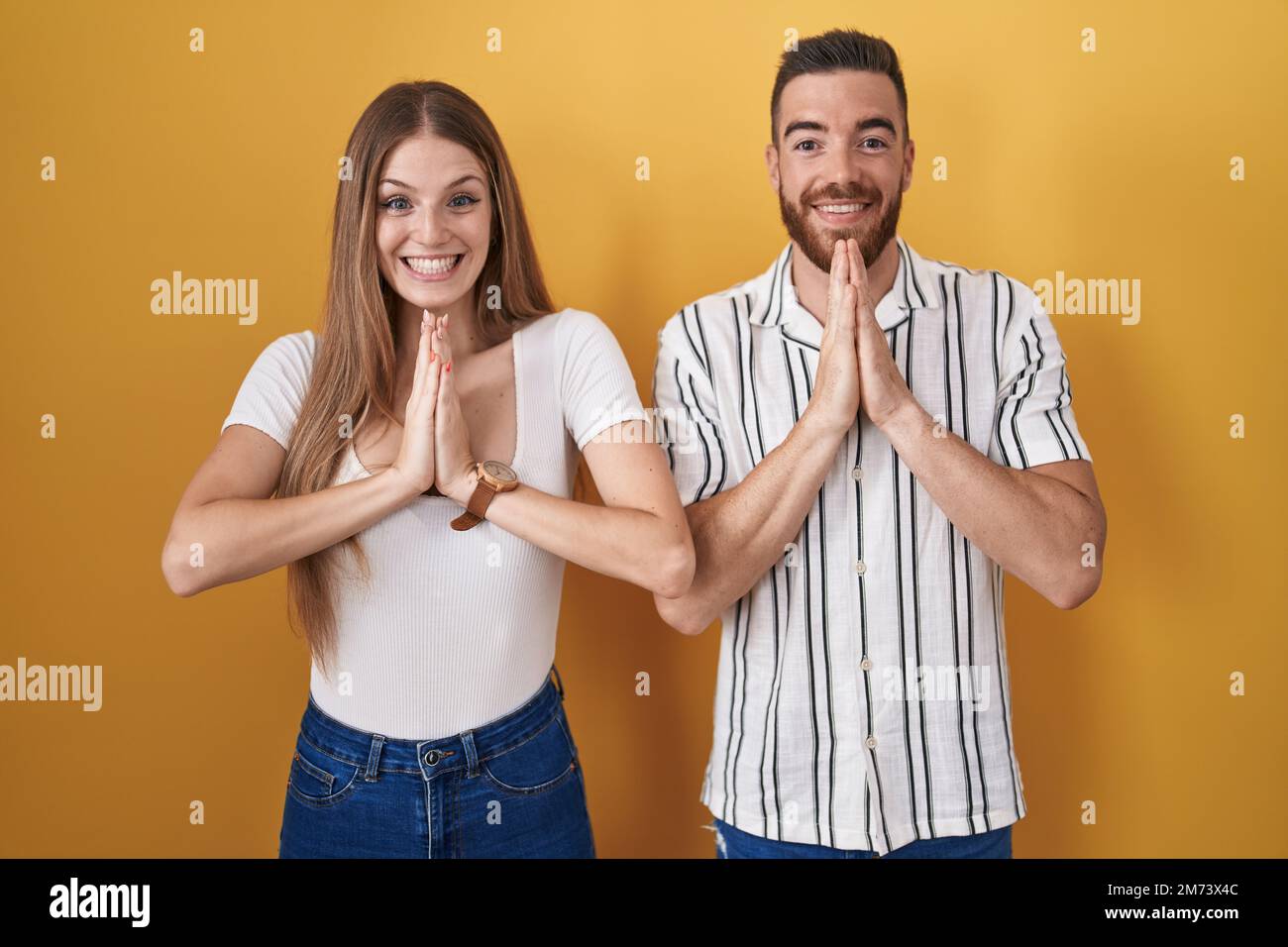 Young couple standing over yellow background praying with hands ...