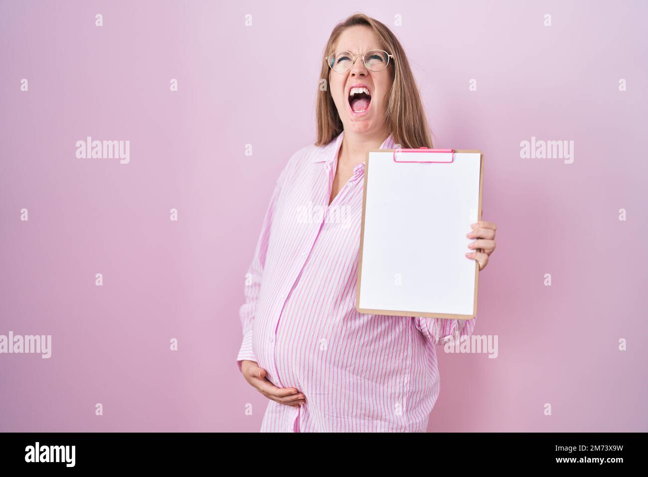 Young pregnant woman holding clipboard angry and mad screaming ...