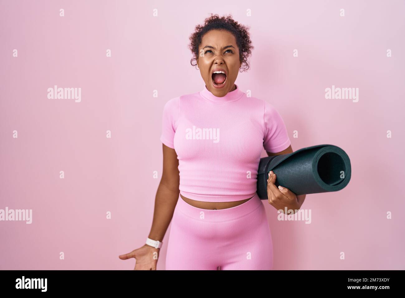 Young hispanic woman with curly hair holding yoga mat over pink ...