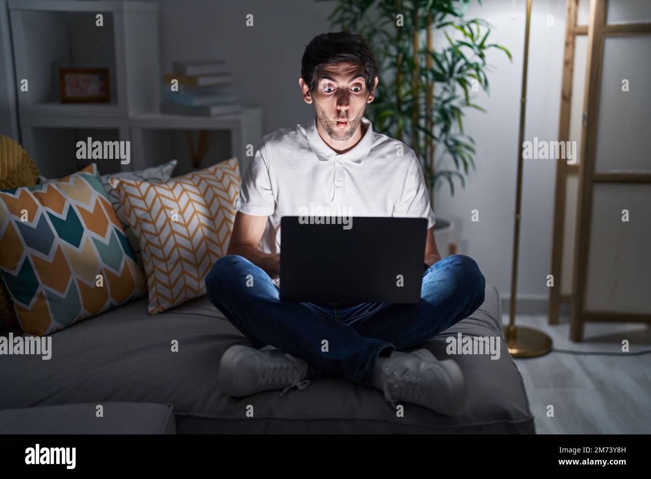 Young hispanic man using laptop at home at night making fish face with ...