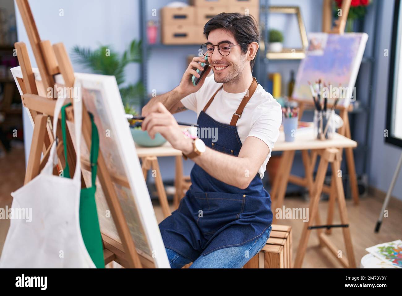 Young hispanic man artist talking on smartphone drawing at art studio ...